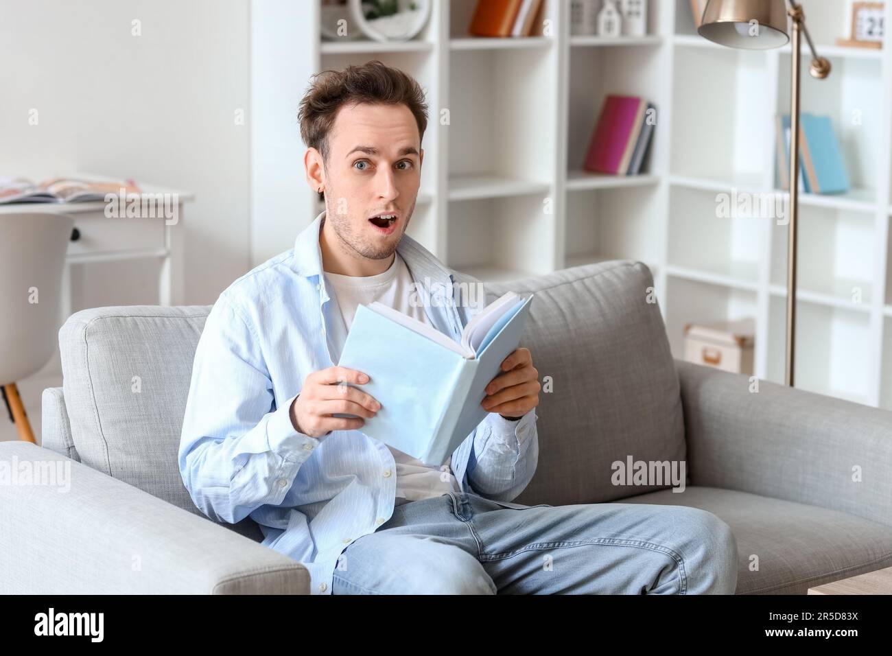 Shocked young man reading book on sofa at home Stock Photo - Alamy