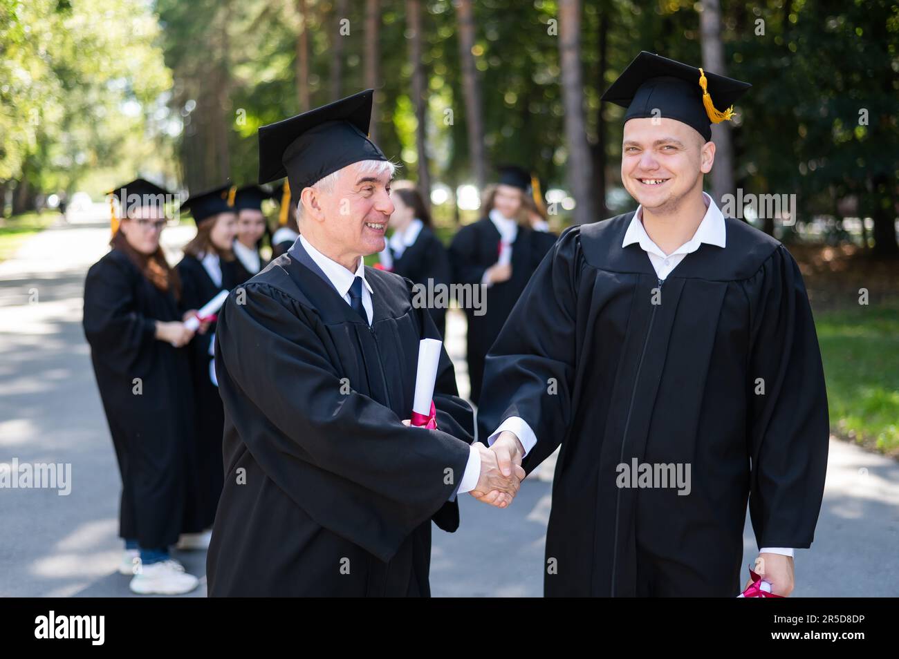 A group of graduates in robes outdoors. An elderly man and a young guy ...