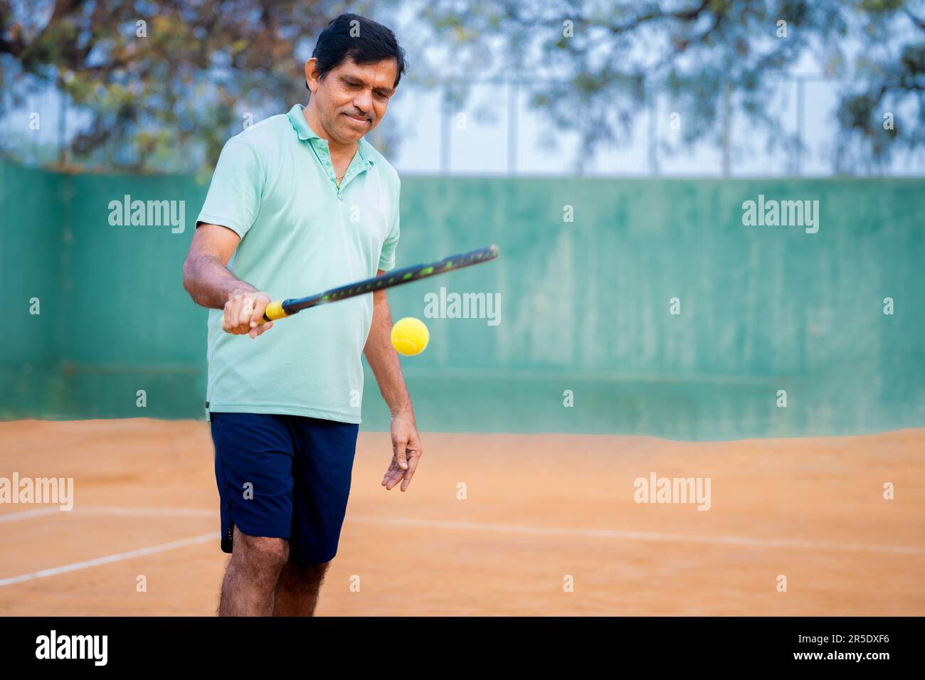 indian senior man practicing tennis by tapping on ground at court ...