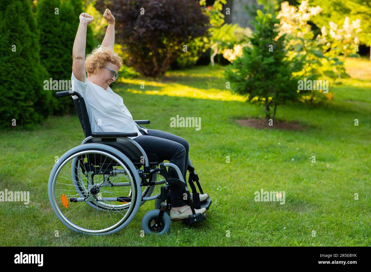 Elderly woman stretches her arms up while sitting in a wheelchair on a ...