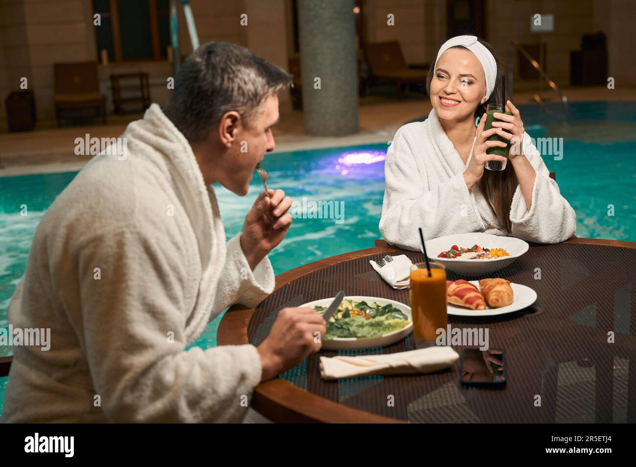 Mature couple in bathrobes having lunch by swimming pool Stock Photo ...