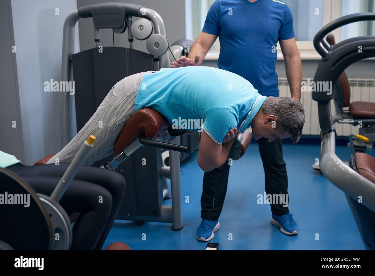 Guy working out on gym equipment supported by fitness trainer Stock ...