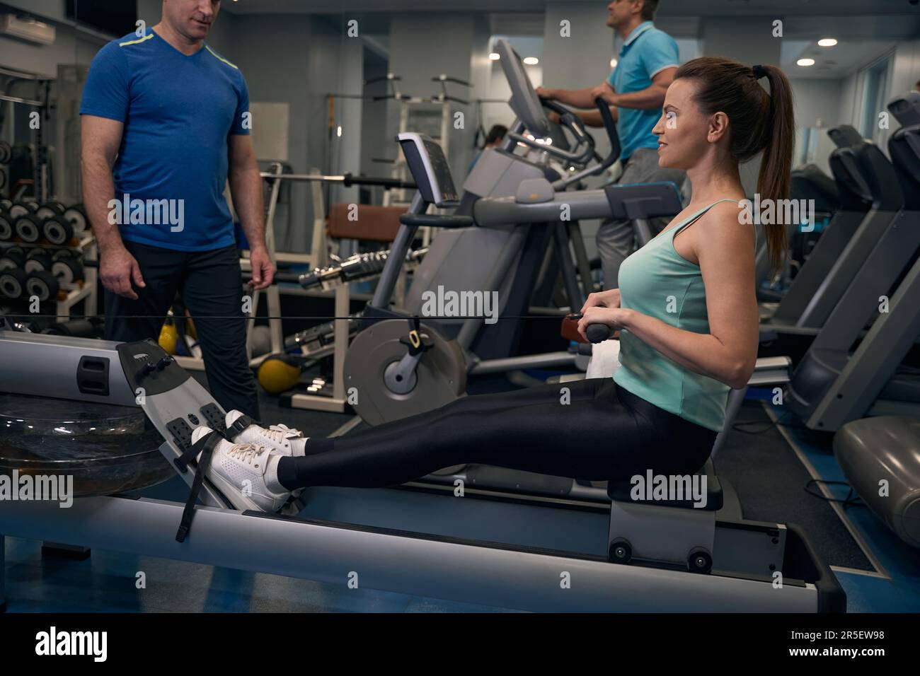 Joyous female working out on gym equipment supervised by trainer Stock ...