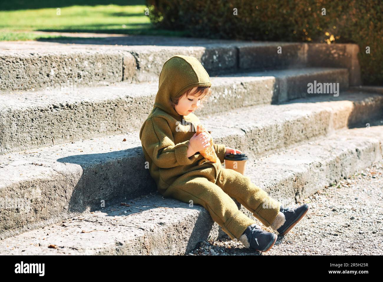 Funny little kid eating croissant and drinking from takeaway coffe cup ...