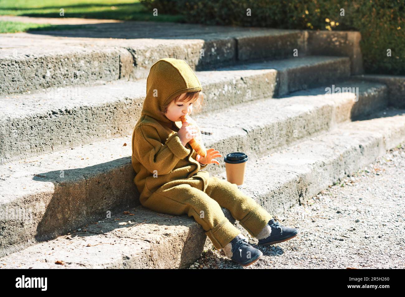 Funny little kid eating croissant and drinking from takeaway coffe cup ...