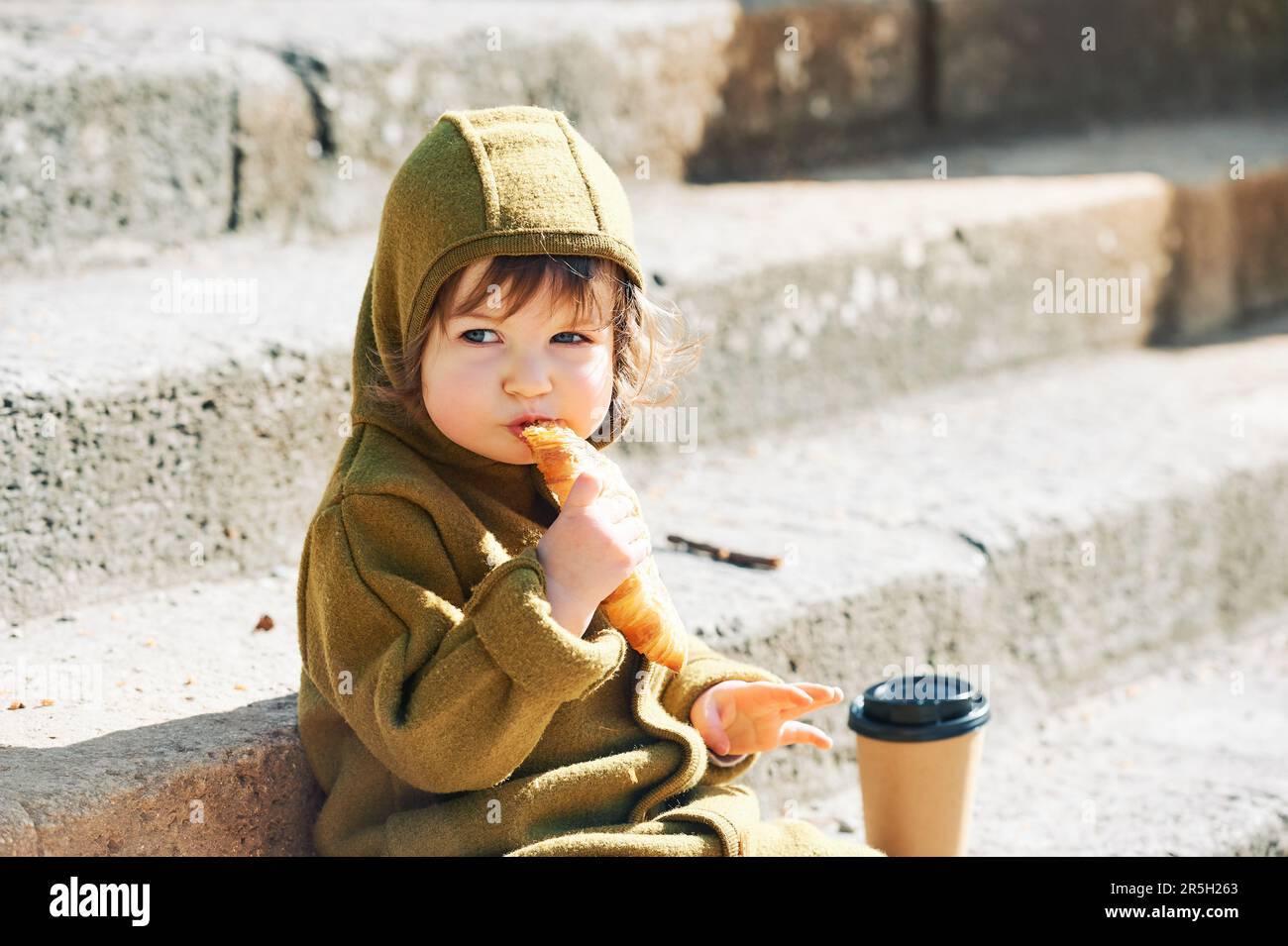 Funny little kid eating croissant and drinking from takeaway coffe cup ...