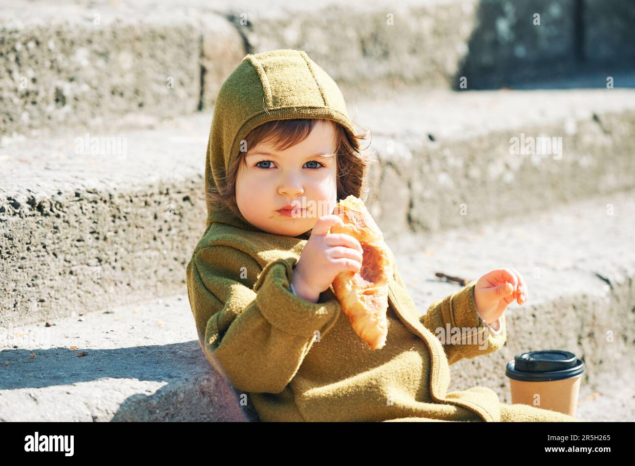 Funny little kid eating croissant and drinking from takeaway coffe cup ...