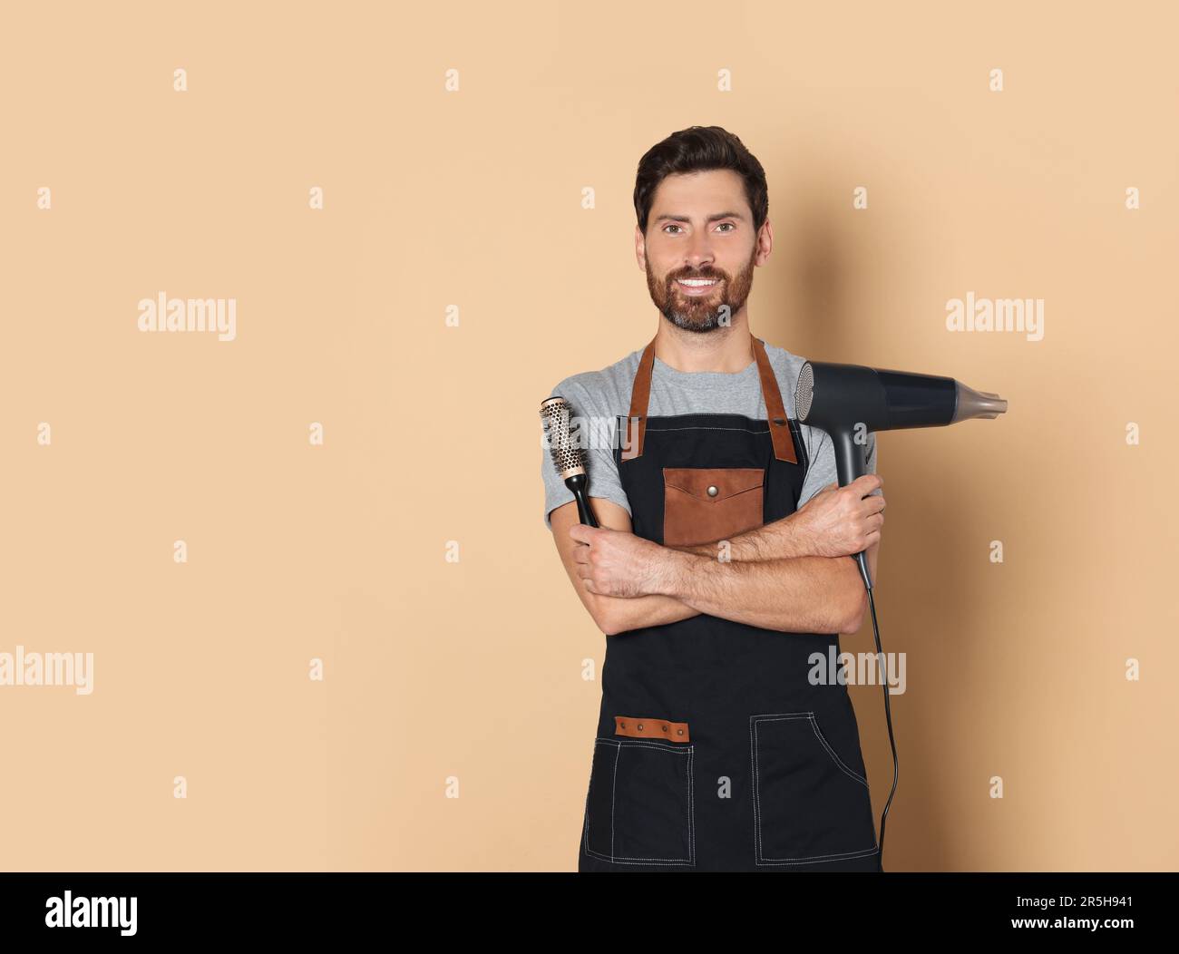 Smiling hairdresser in apron holding dryer and brush on light brown ...