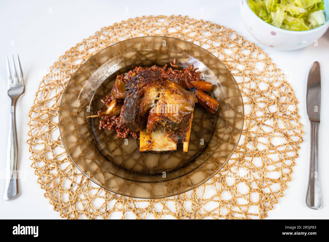 Baked beef rib with red rice on brown plate, cutlery, bamboo pad, salad ...