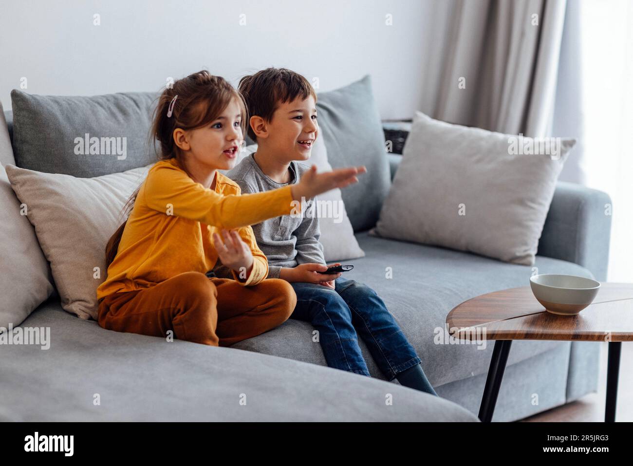Two little kids are sitting on grey sofa and watching television. Cute ...