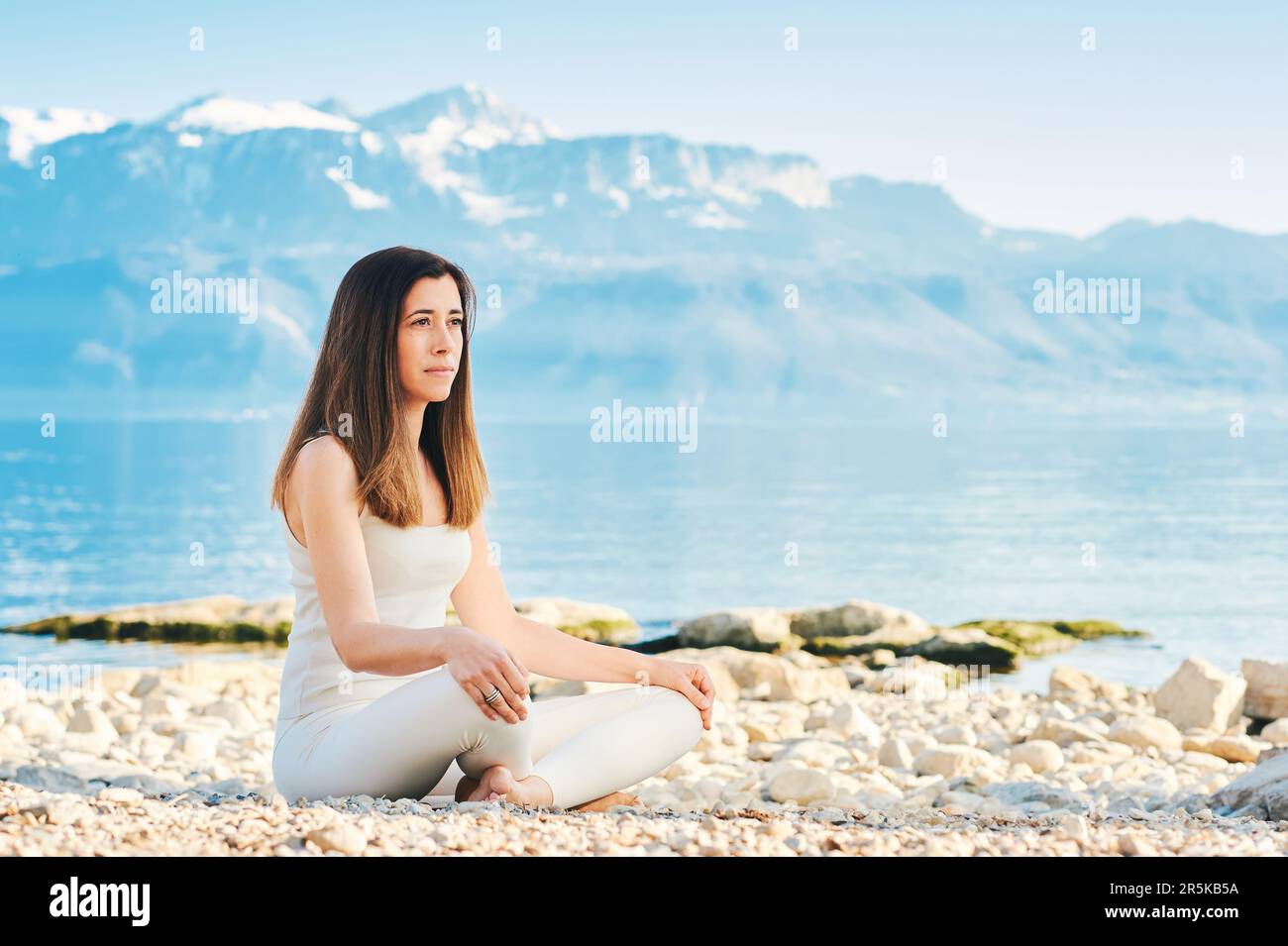 Beautiful spiritual woman meditating by the lake, wearing beige clothes ...