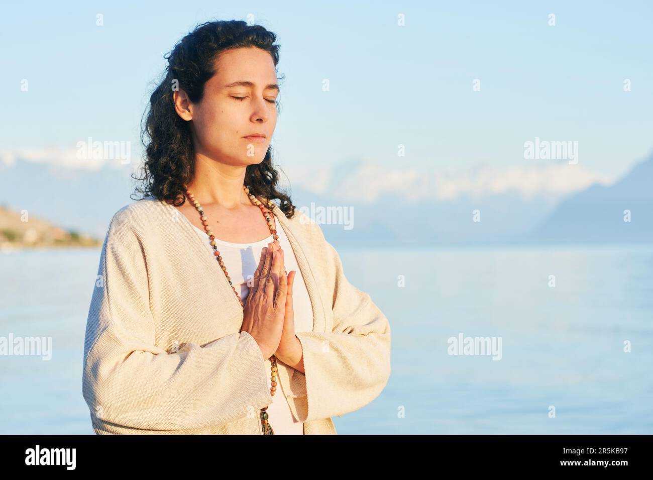 Beautiful spiritual woman meditating by the lake, wearing beige clothes ...