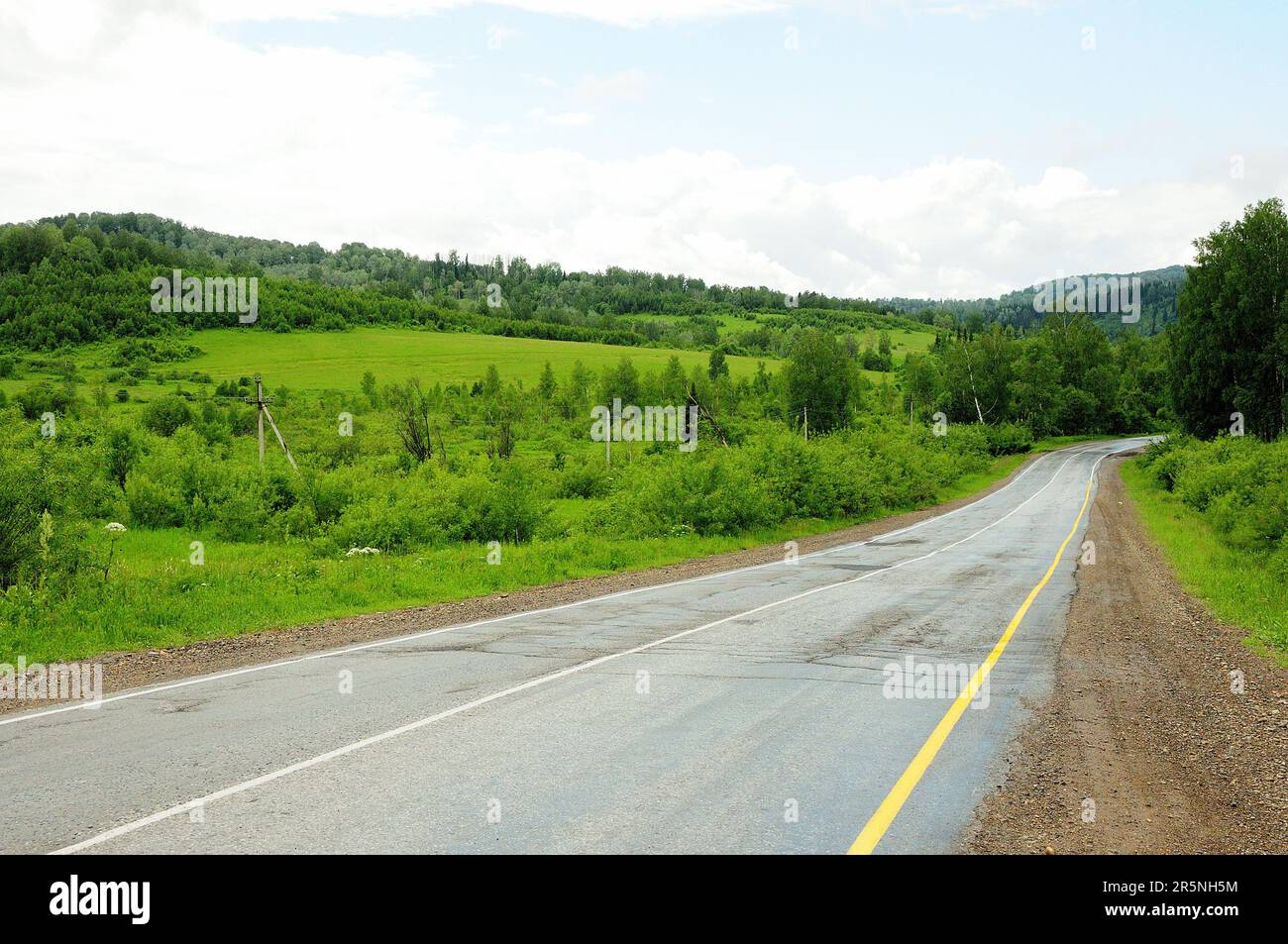 A two-lane asphalt road curves through a beautiful hilly valley under a ...