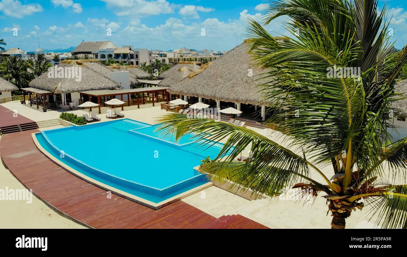 Aerial view of luxury hotel with straw roof villas and pools in tropical resort and palm trees ...