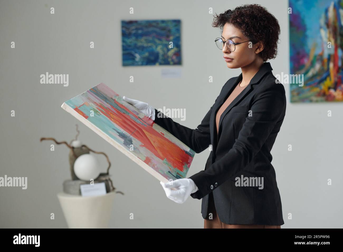 Young gallery owner in white gloves holding painting in her hands while ...