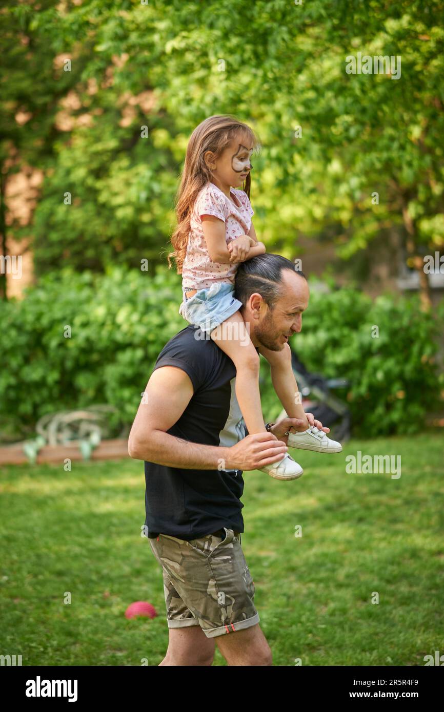 A cheerful father carrying his daughter on his shoulders, laughing and looking up at the sky ...