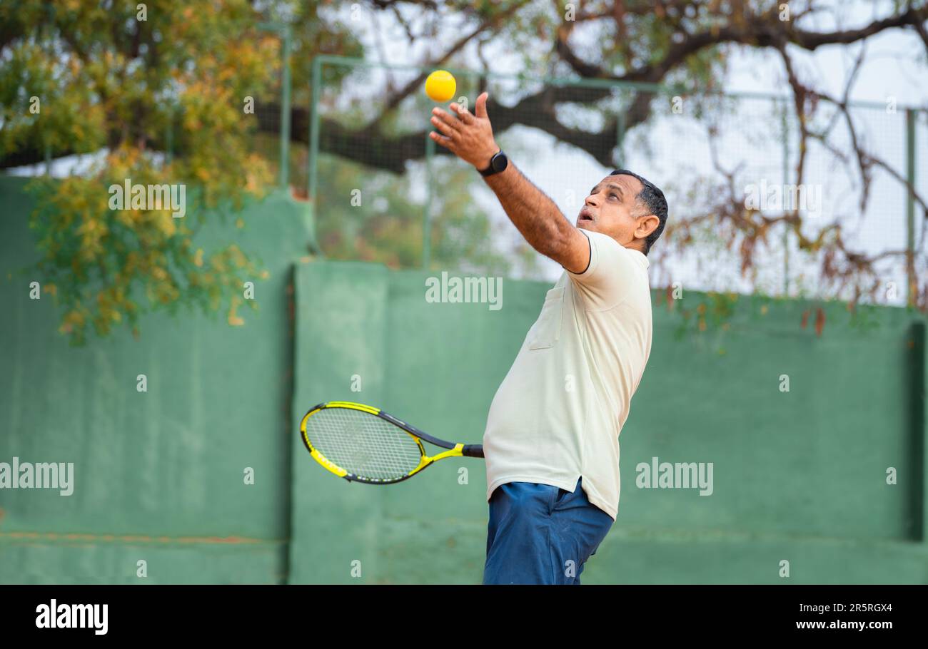 Indian senior man servicing by throwing ball at tennis court while ...