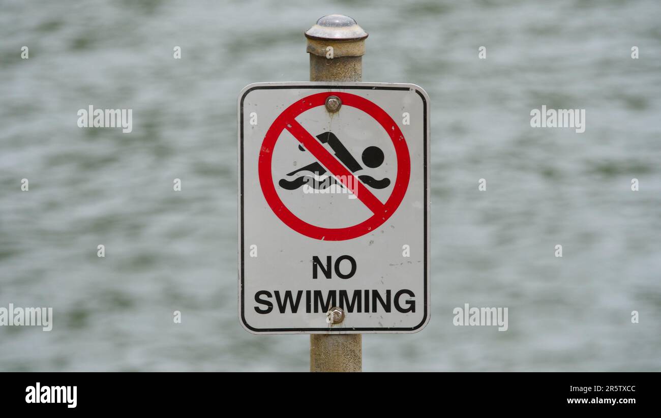 A close-up of a sign warning against swimming, beside a body of water ...