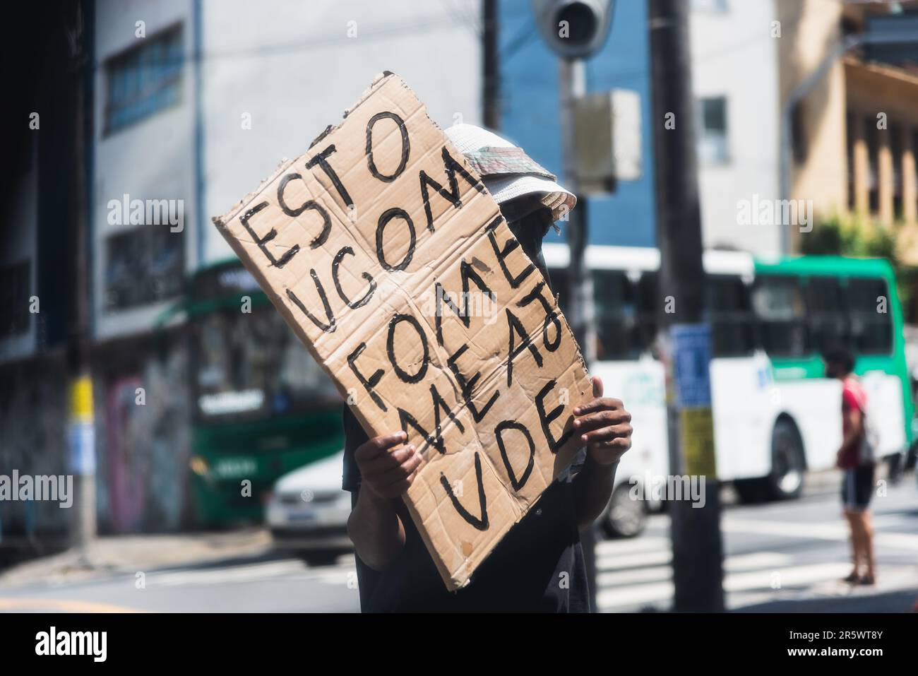 A homeless individual stands in the street, holding a cardboard sign ...
