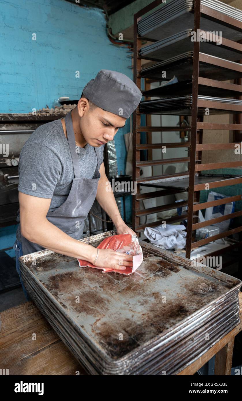 A young Hispanic baker is cleaning a tray with butter before baking. He ...