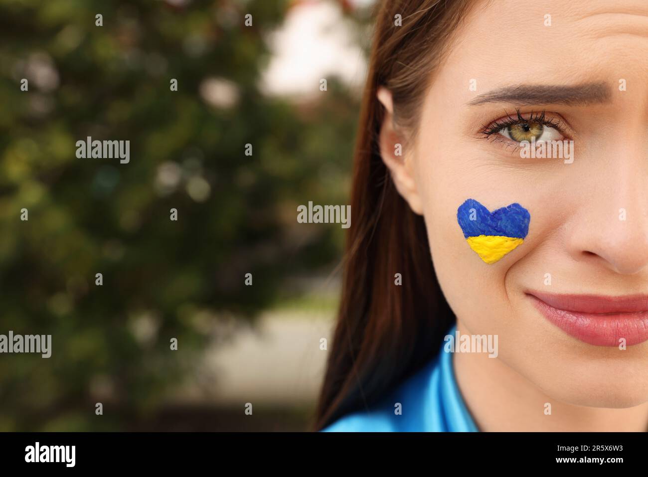 Sad young woman with drawing of Ukrainian flag on face outdoors ...