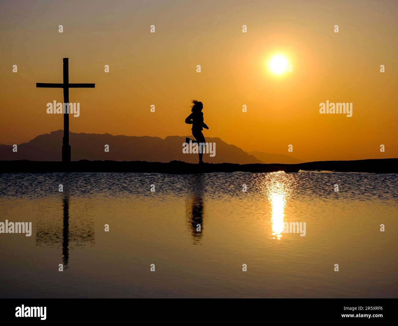 Silhouette of a runner with summit cross at sunset, reflection in the ...