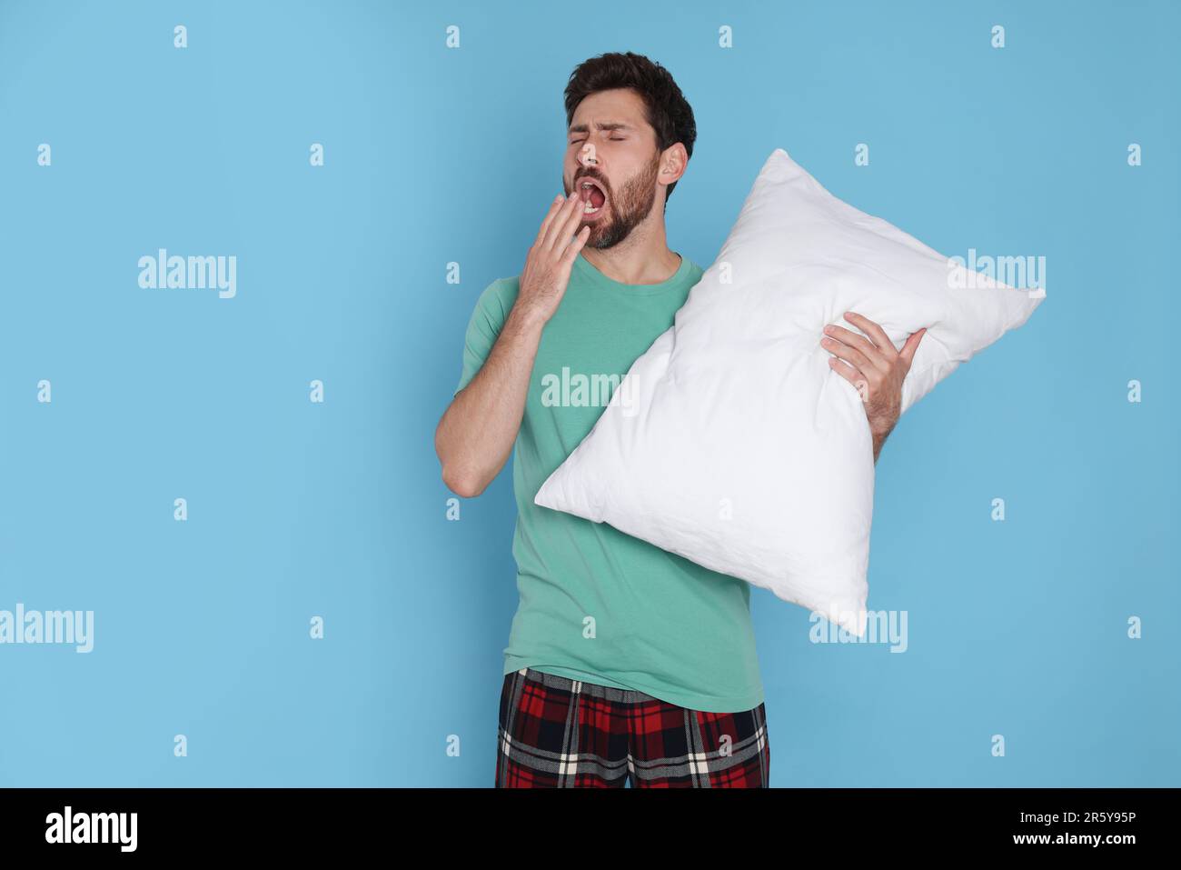 Tired man with pillow yawns on light blue background, space for text ...