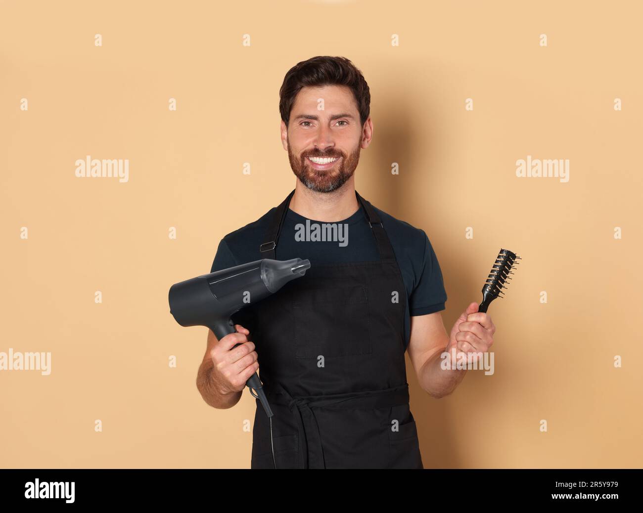 Smiling hairdresser in apron holding dryer and brush on light brown ...