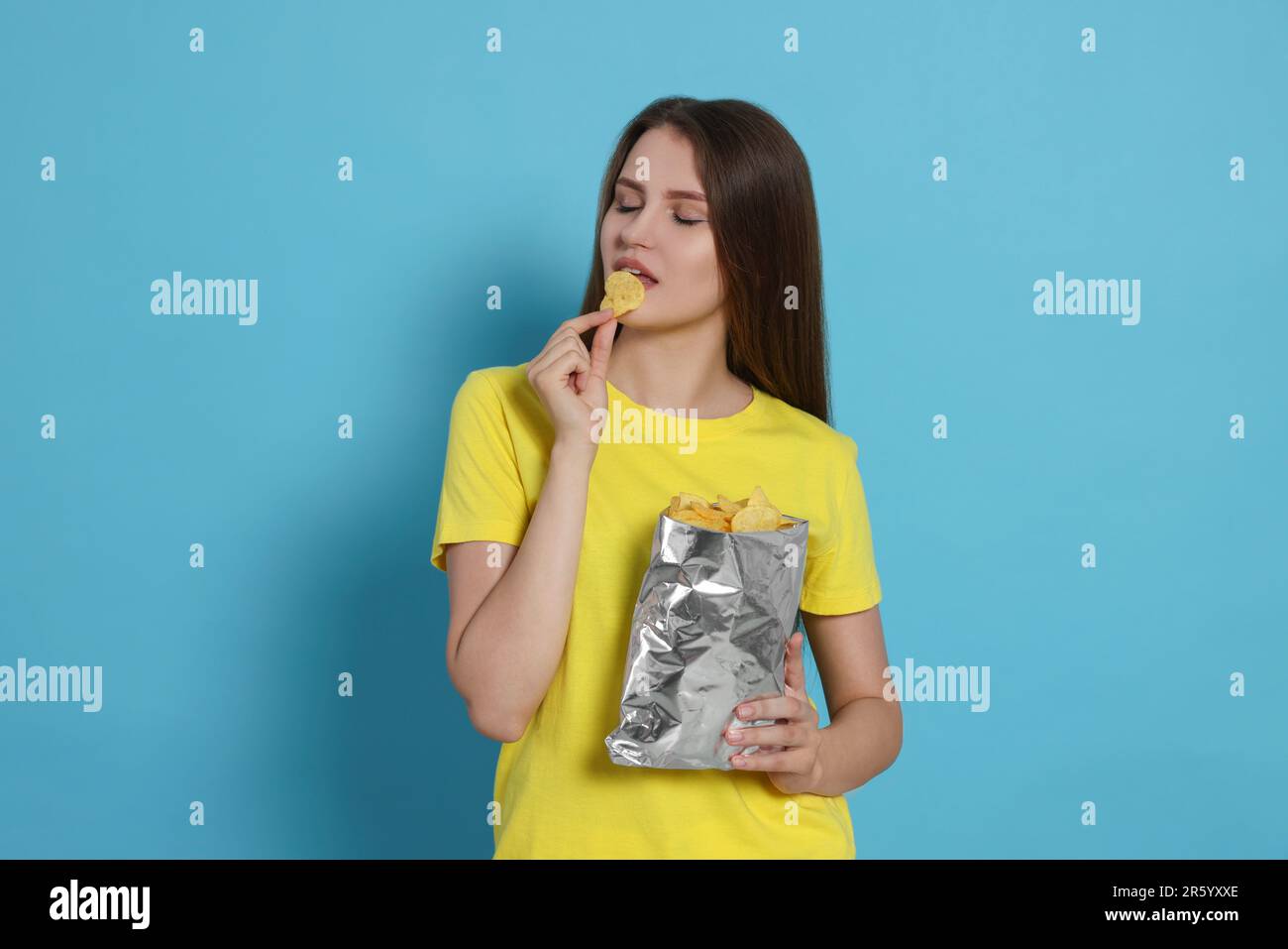 Pretty young woman eating tasty potato chips on light blue background ...