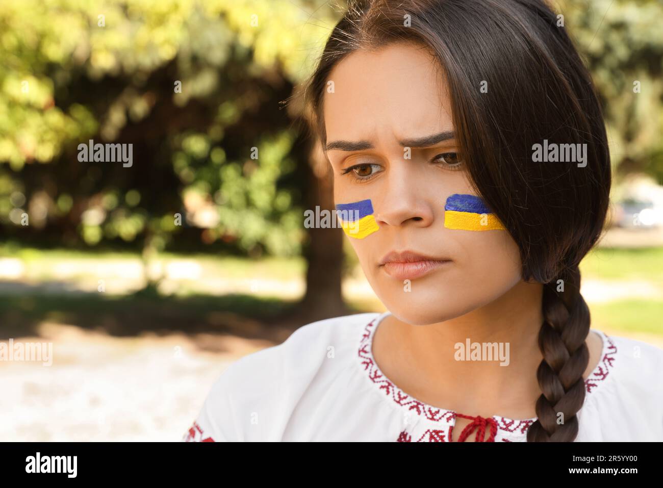 Sad young woman with drawings of Ukrainian flag on face in park, space ...