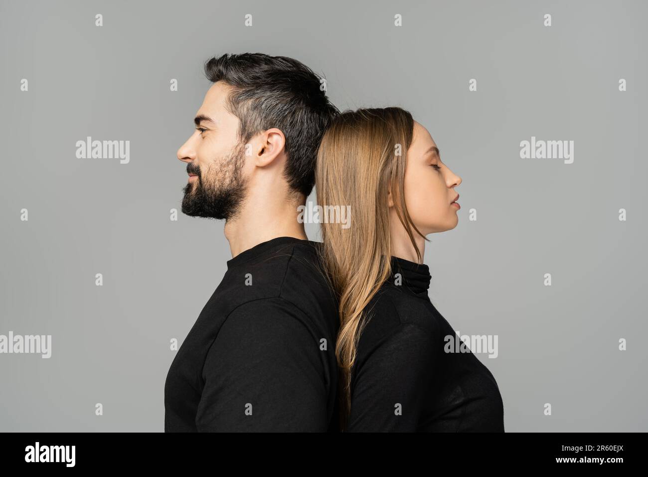 Side view of relaxed and fair haired woman standing back to back with bearded husband in black t ...
