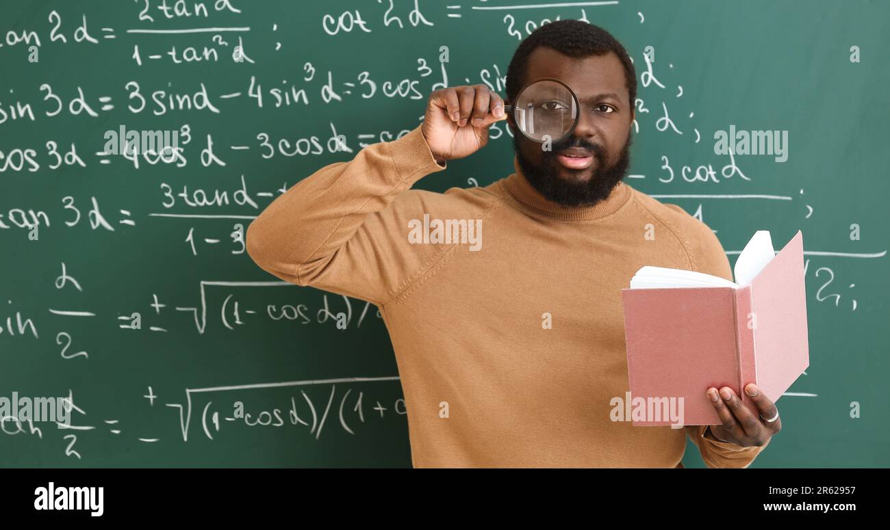 African-American math teacher with book and magnifier near blackboard ...