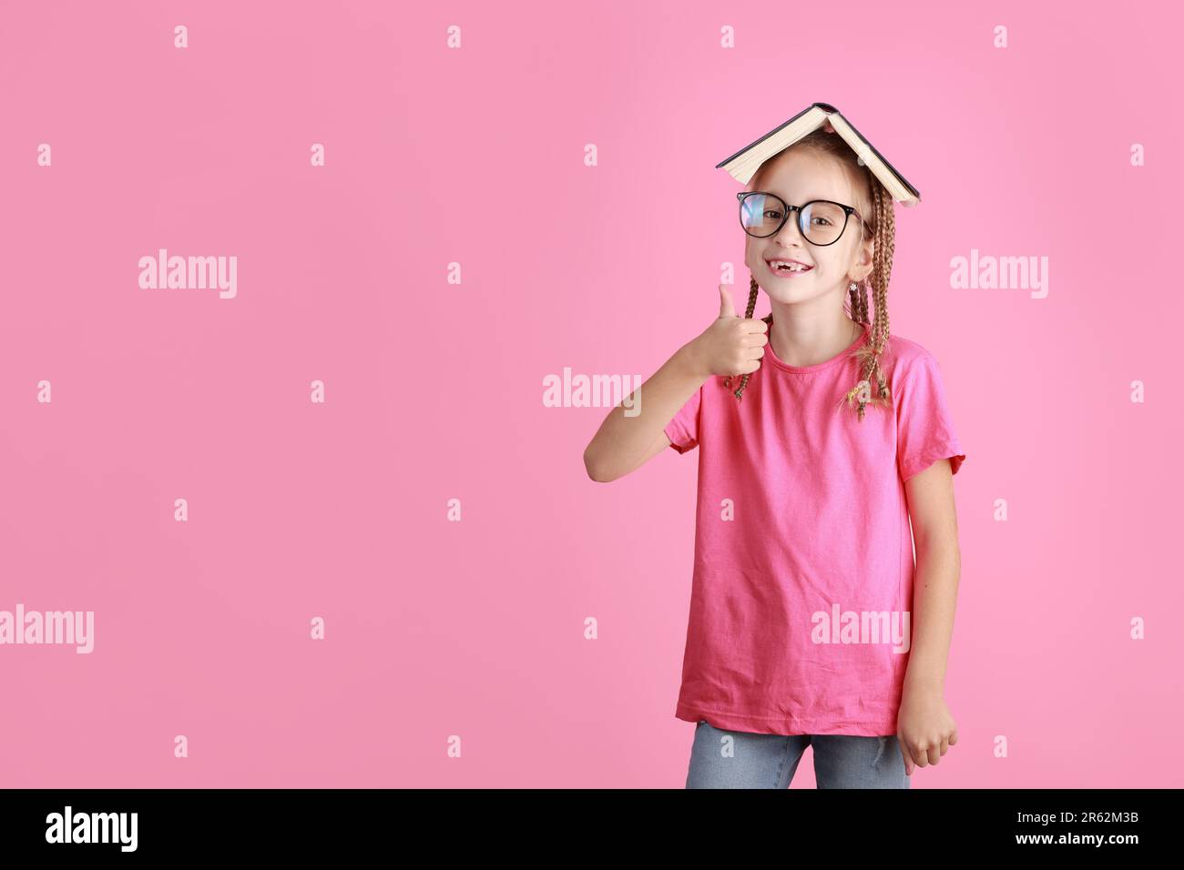 Back to school concept. Smiling pretty little school girl in glasses is holding book over head ...