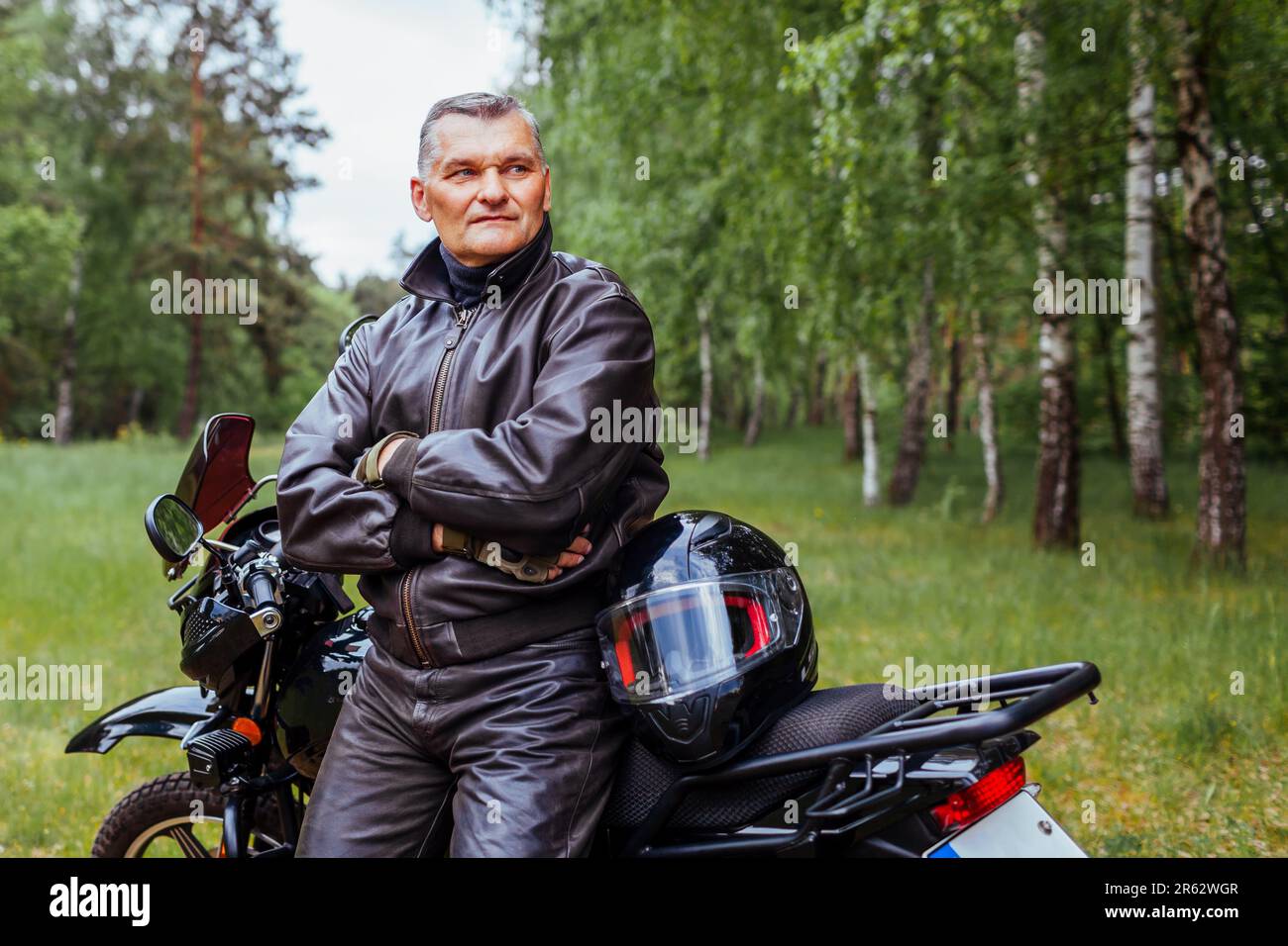Elderly biker leaning on motobike in summer forest. Man in leather ...