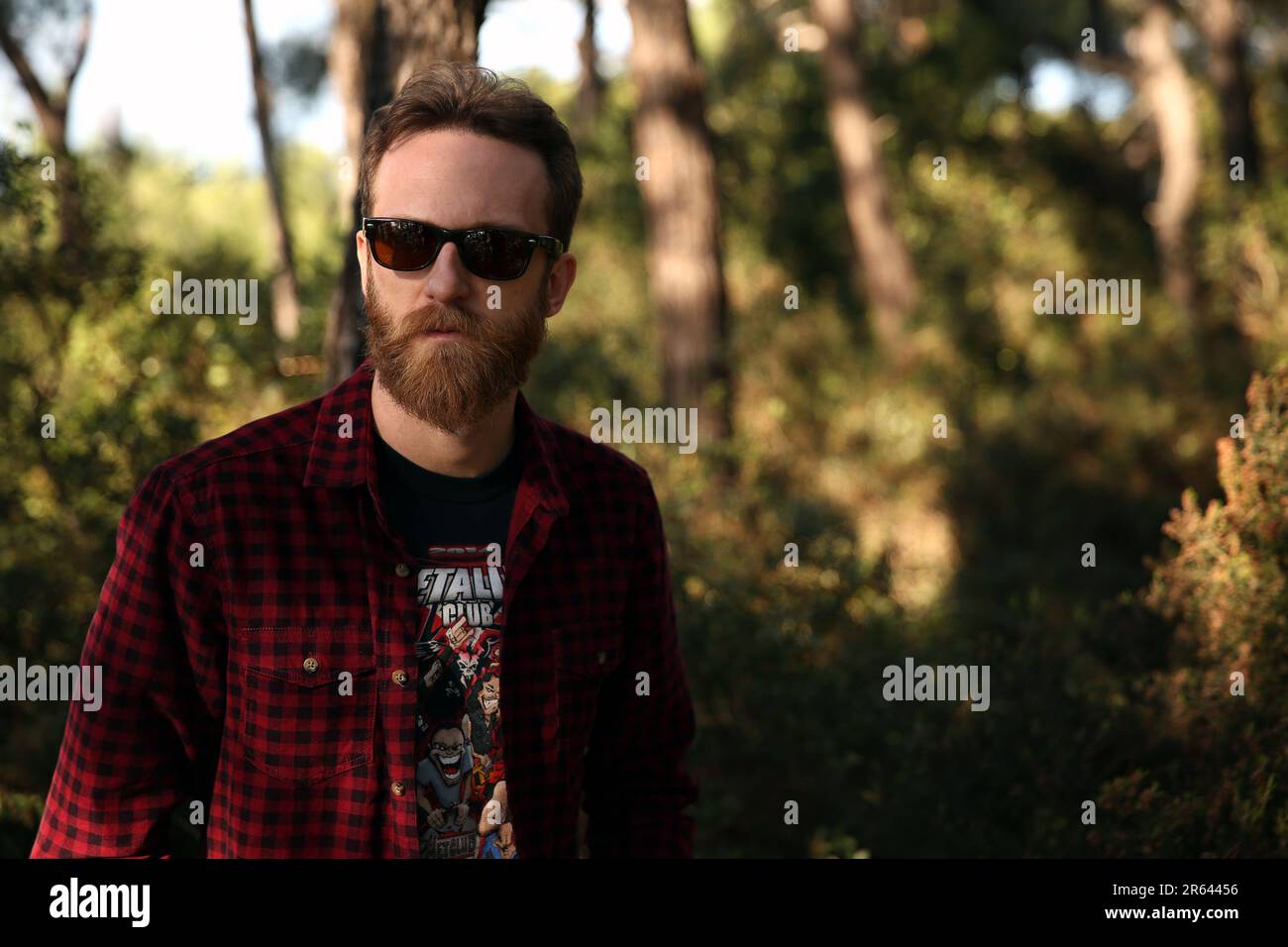 Bearded man with sunglasses and a red shirt posing in front of a blurry ...