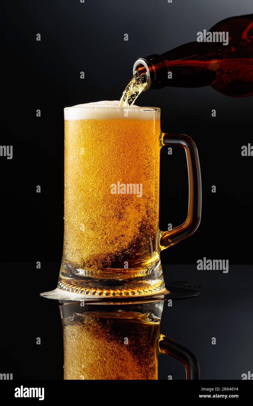 Pouring beer from a bottle into a mug. Beer mug on a black reflective ...
