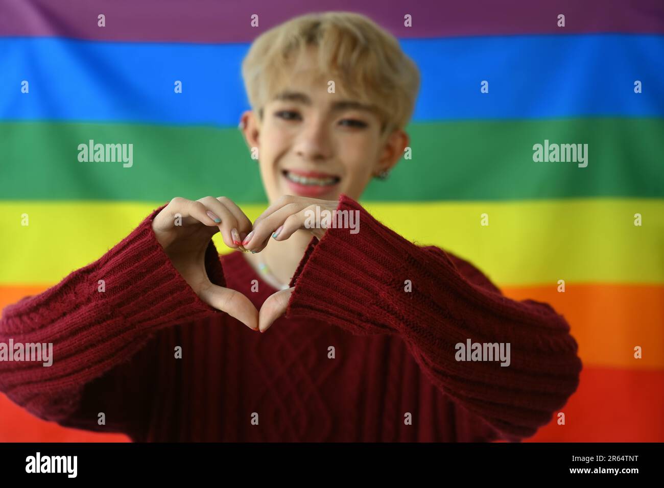 Cheerful young Asian gay man making heart with hands over rainbow pride ...