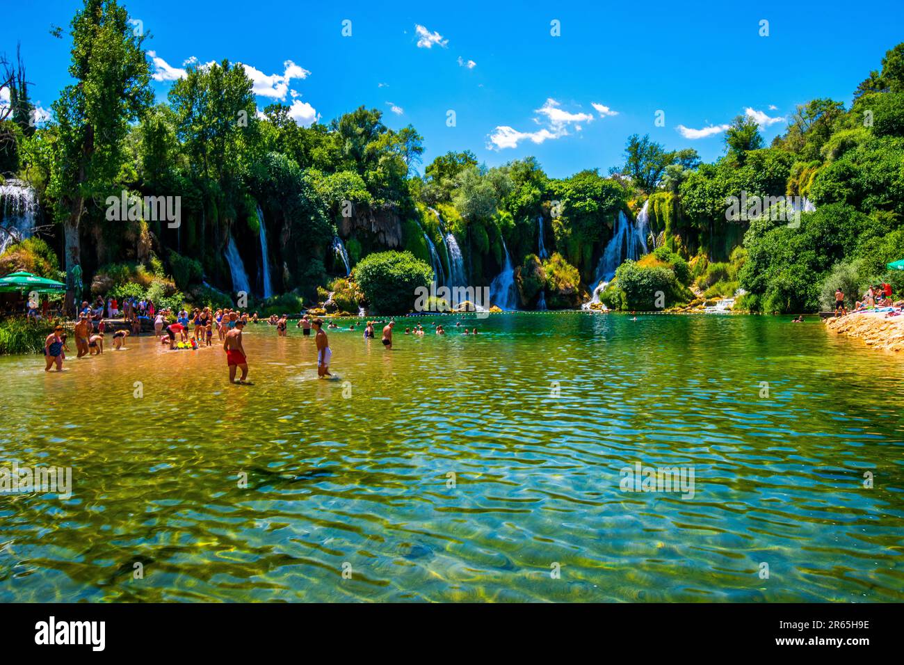 A group of people stand on the edge of a river with a waterfall in the ...