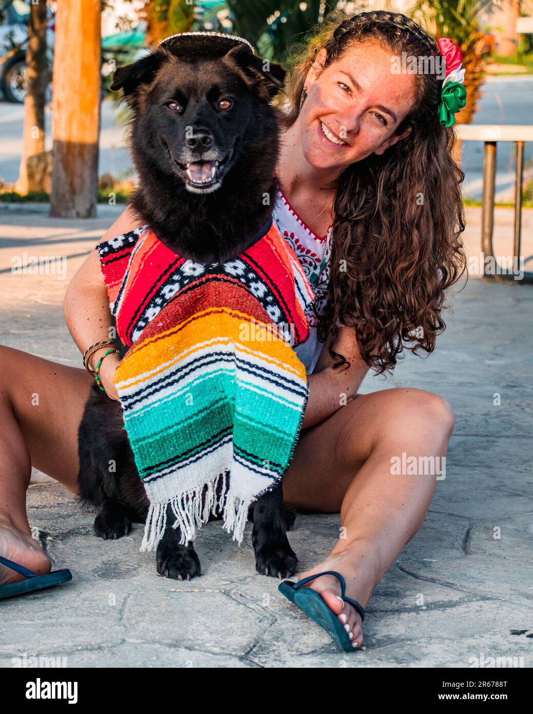 A cheerful young woman sits on the ground in a park, petting and ...