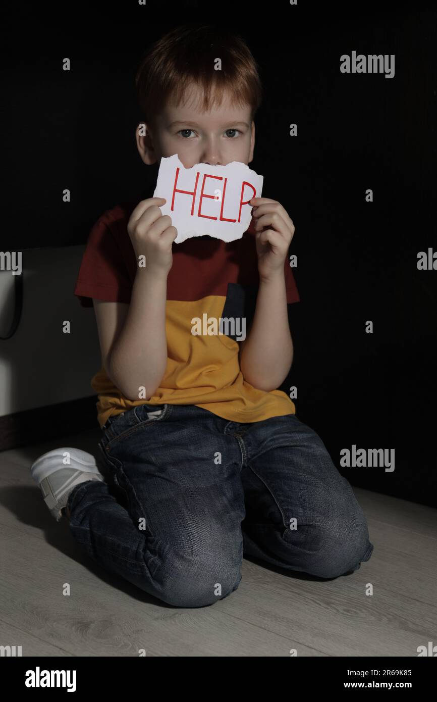 Scared little boy holding piece of paper with word Help under table on ...