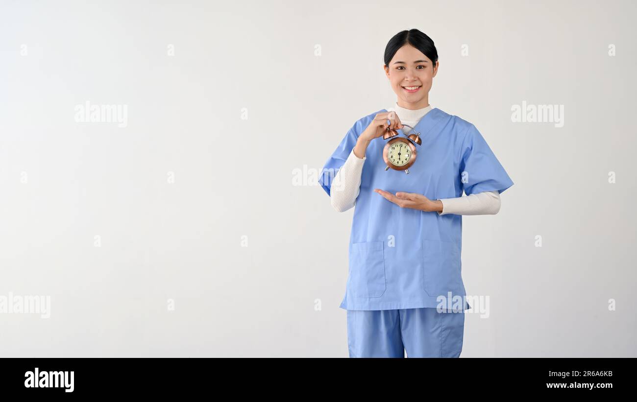 A smiling Asian female doctor in scrubs holds a vintage alarm clock ...