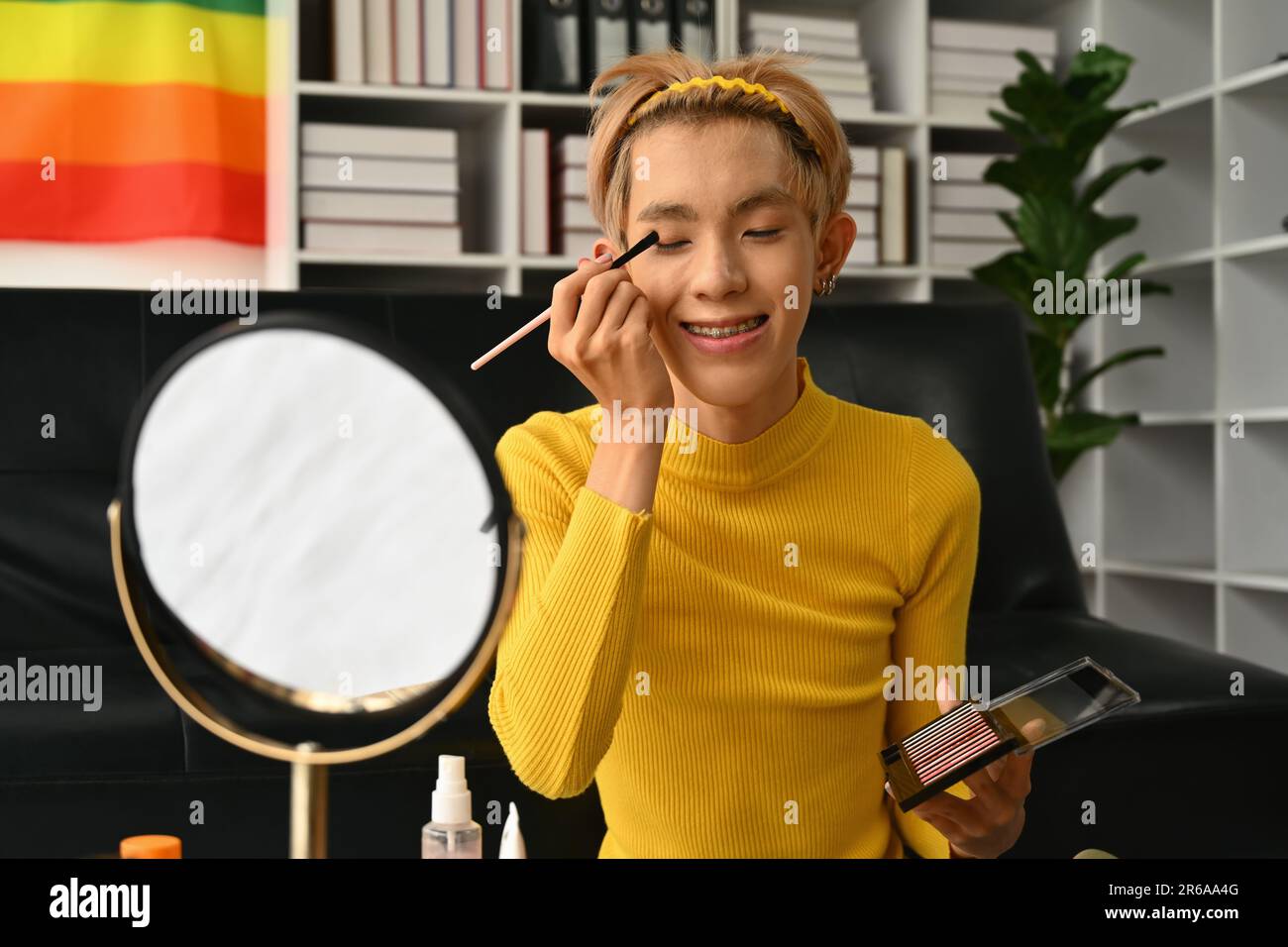 Happy young gay man applying eye shadow with applicator near mirror ...
