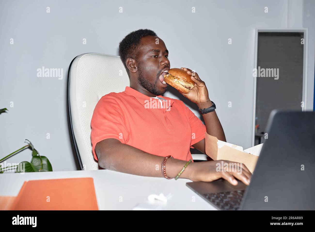 Side view portrait of black man eating burger at workplace and using ...