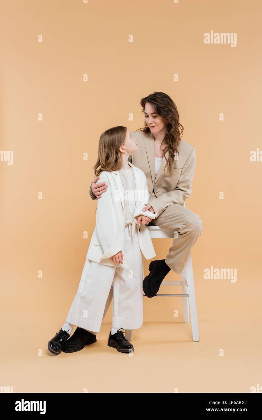 stylish mother and daughter in suits, woman sitting on high chair and ...