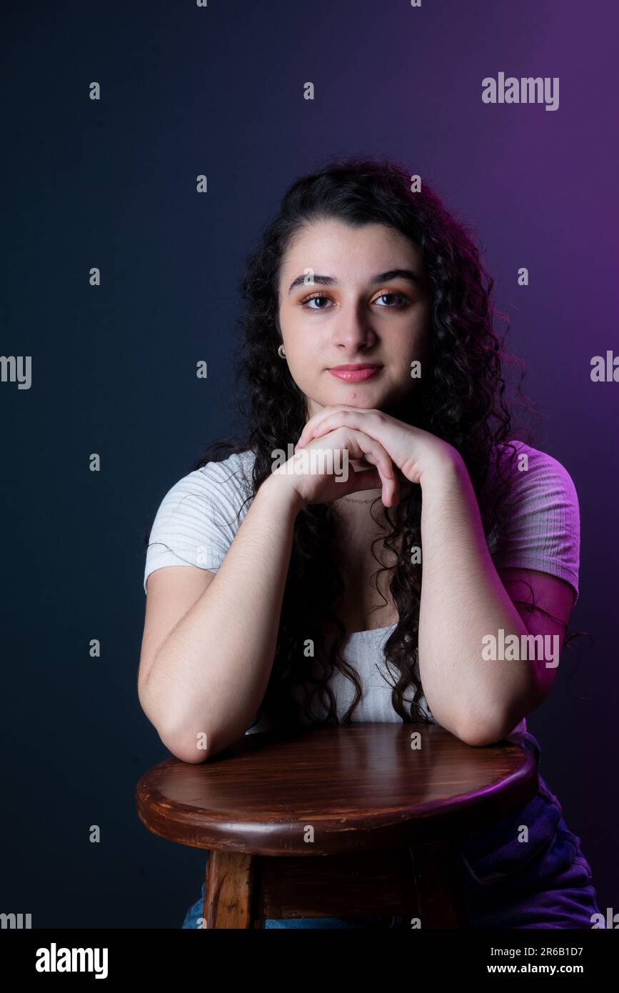 A young female adult is seated on a stool with her hands clasped ...