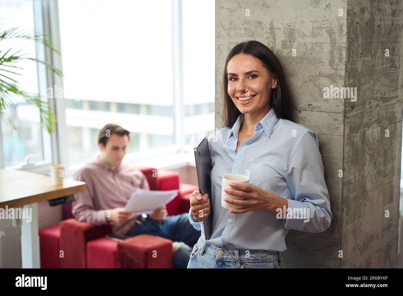 Joyful company employee with teacup and laptop leaning against pillar ...