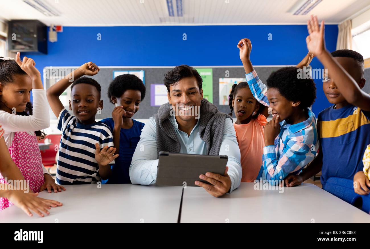 Diverse male teacher with tablet and elementary schoolchildren raising ...