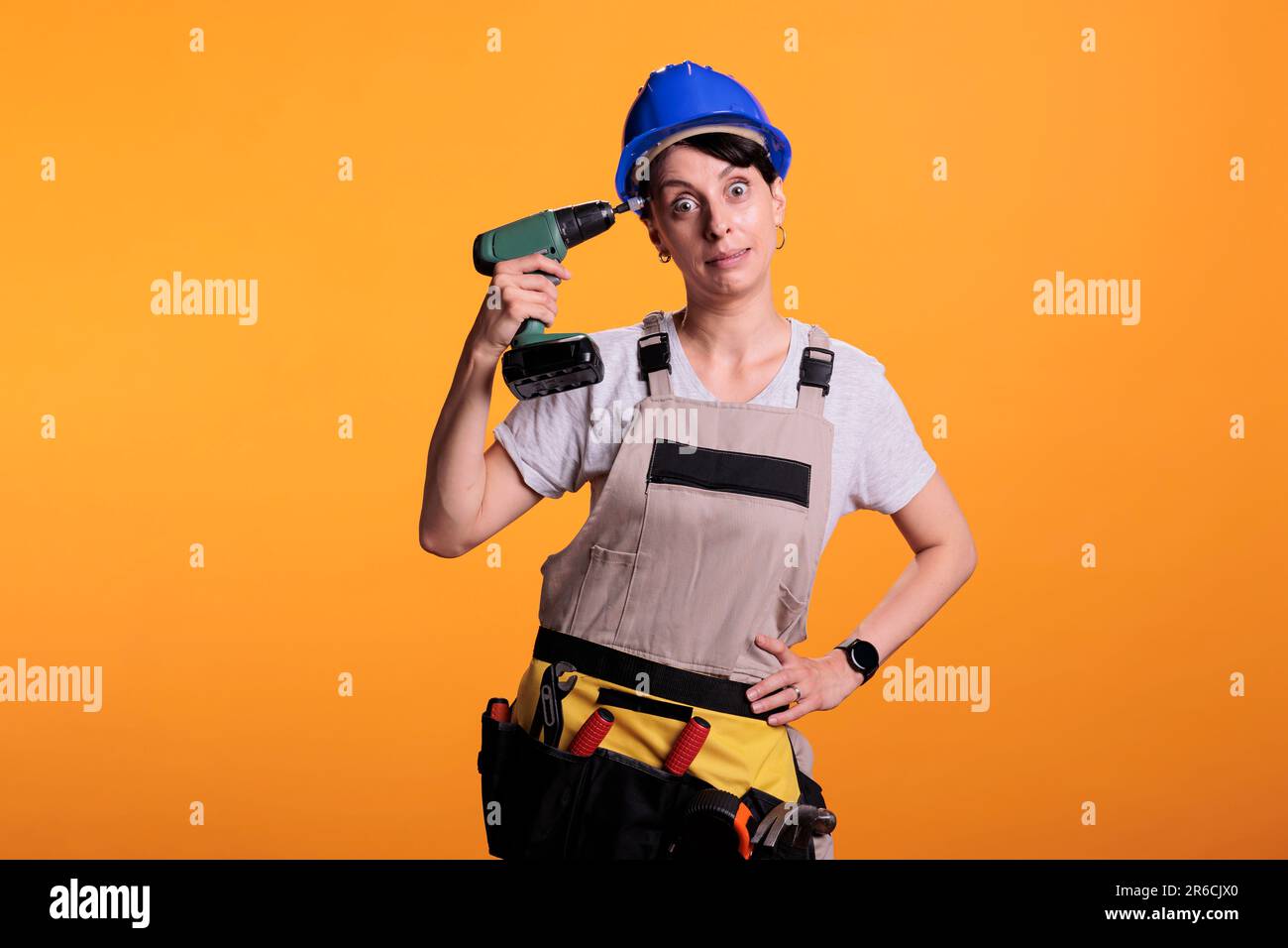 Female builder in hard hat holds electric drill against her head, going ...