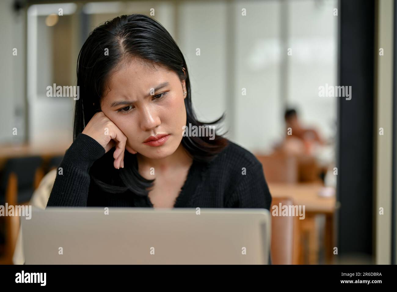 Puzzled and stressed young Asian woman is looking down at her laptop ...