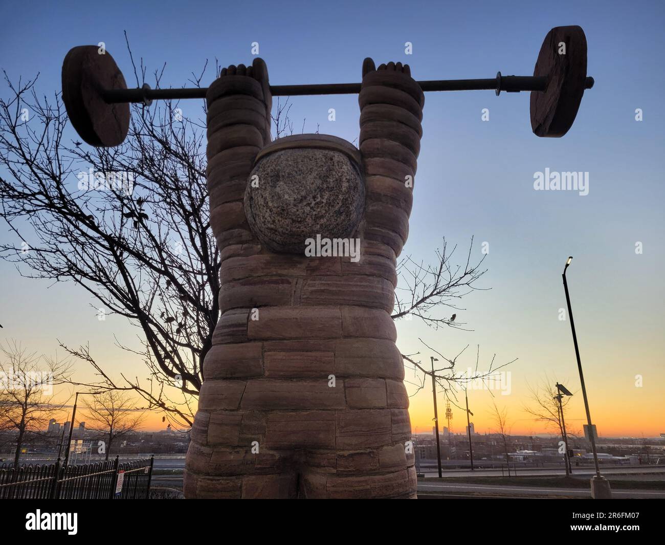 A large sculpture is situated beside a riverbank, with two weights held ...